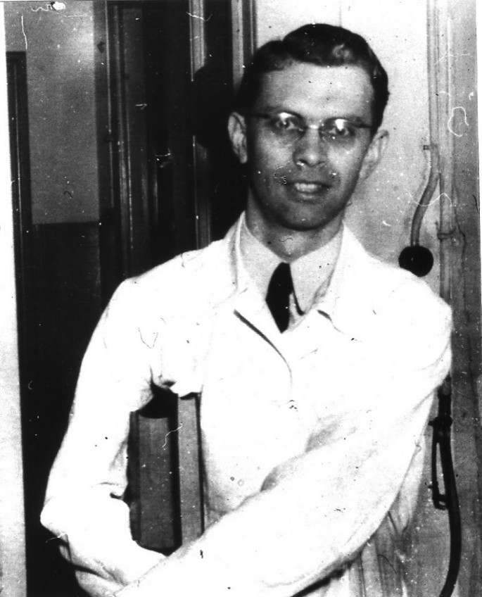 A photo of a man who stands at the entrance to a lecture hall, wearing glasses. He holds books under his arms. The partially open door of the hall features a curved metallic handle.