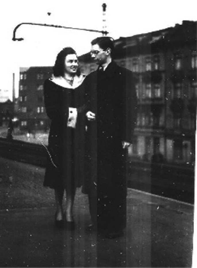 A photo of a man and woman who stand on a road with a street light and buildings in the background. He wears a long coat and glasses, while she wears a midi dress. She gazes at him with a smile on her face.